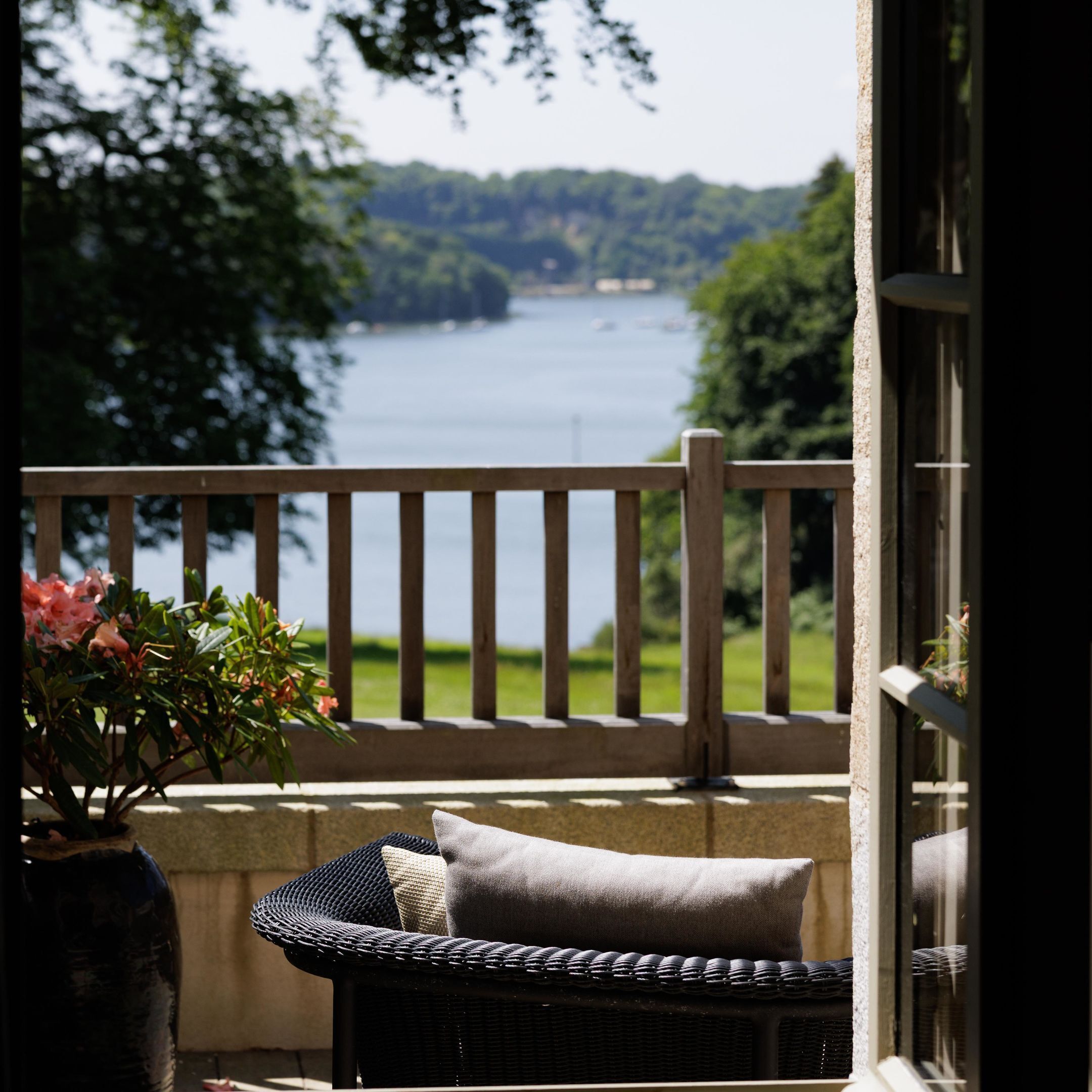 Uma varanda com cadeira de vime e almofadas, com vista para um lago com barcos e vegetação.