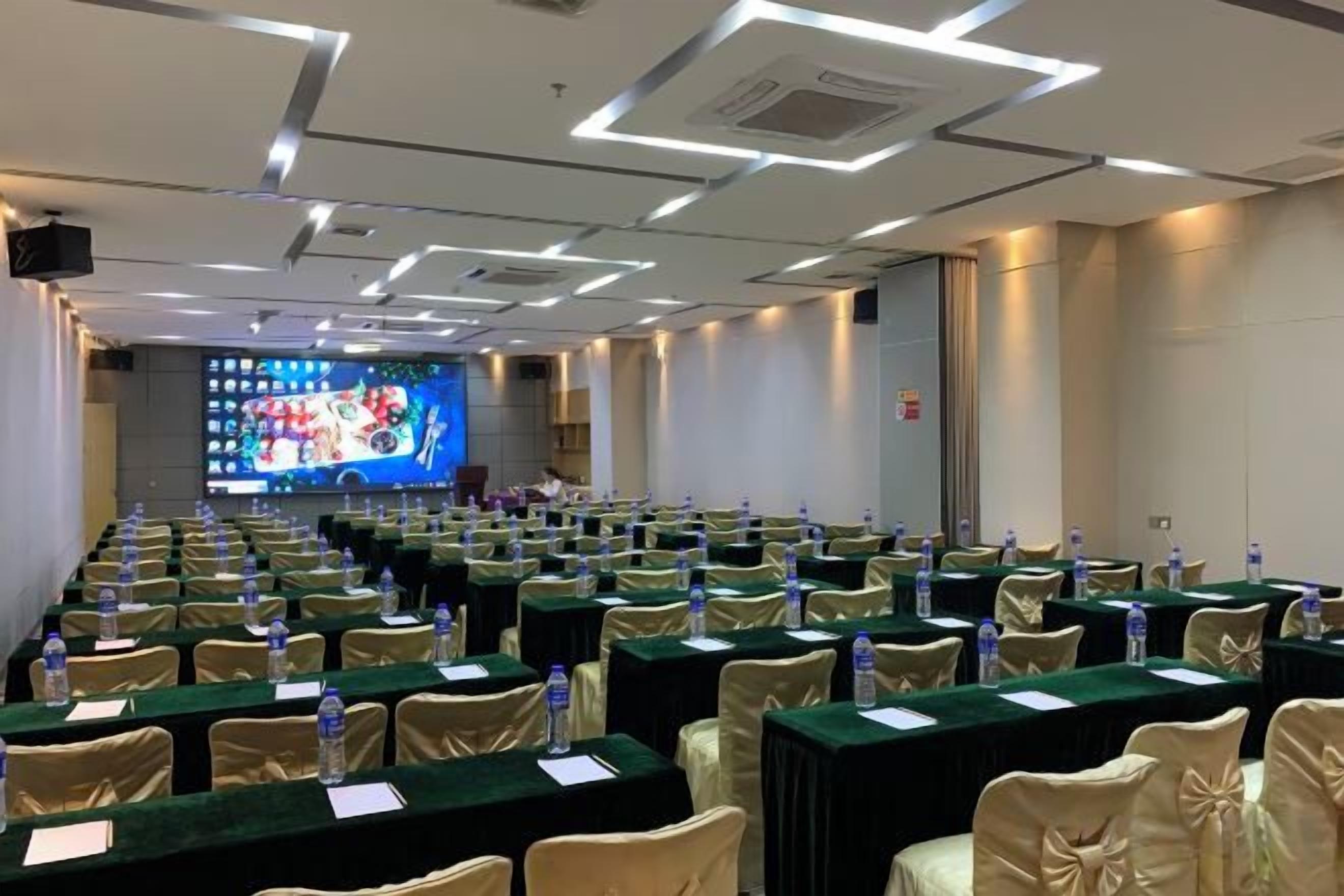 Sala de conferência com fileiras de mesas, cadeiras e uma tela grande na frente.