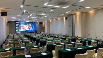 Sala de conferência com fileiras de mesas, cadeiras e uma tela grande na frente.