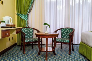 Quarto de hotel com uma mesa redonda de madeira, duas cadeiras verdes com padrão, uma cama, uma mesa de cabeceira com abajur e um tapete estampado.
