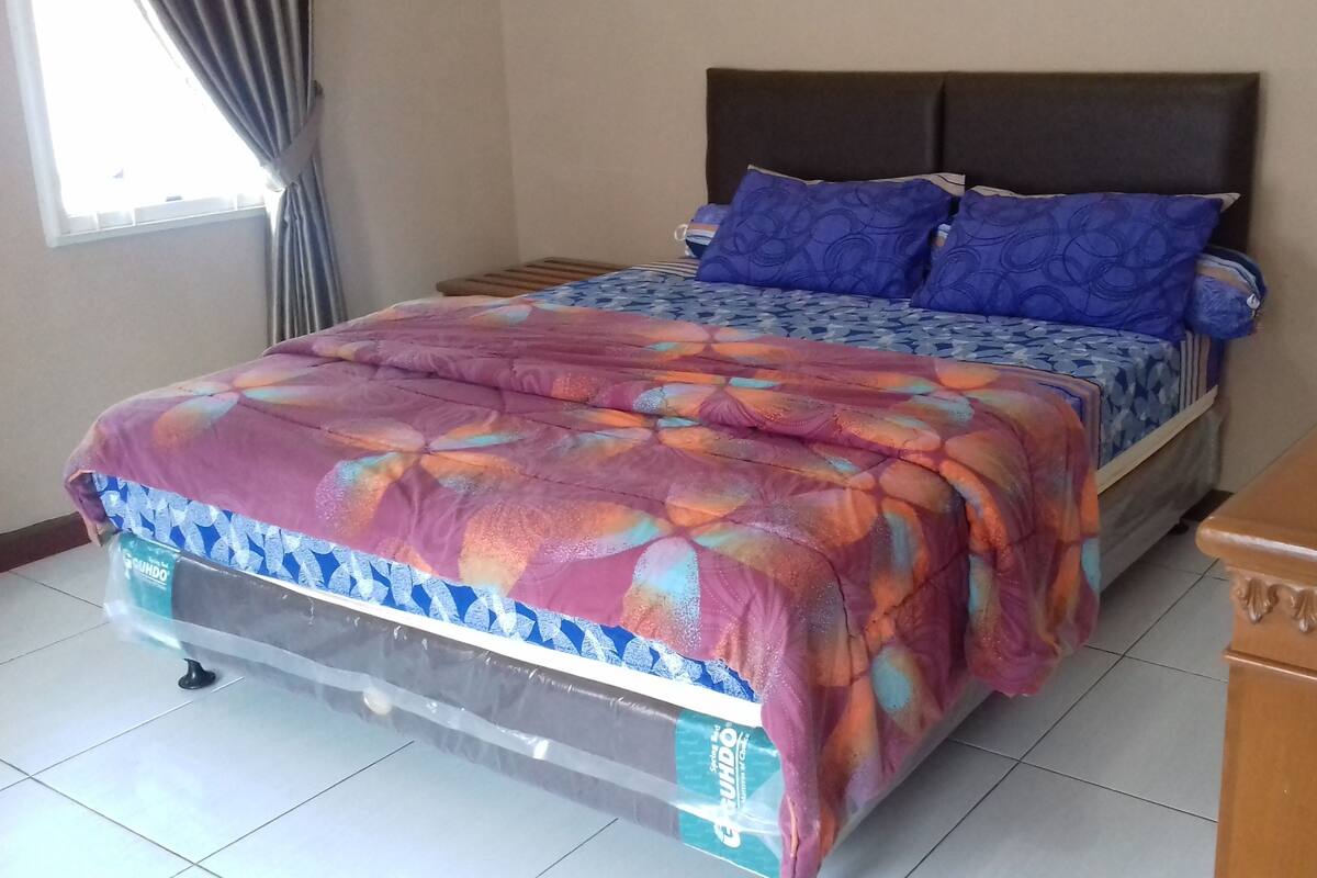 Uma cama bem arrumada com colcha estampada e travesseiros, uma mesinha de cabeceira de madeira e um armário.