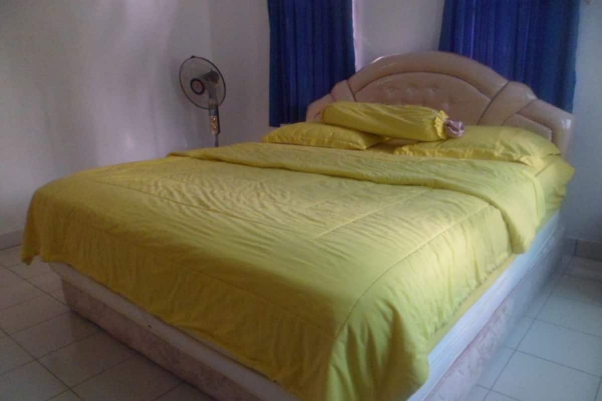Uma cama com colcha e travesseiros amarelos, cabeceira e ventilador fixado na parede.