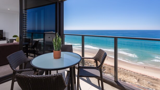 Uma varanda com mesa e cadeiras de vidro, um vaso com planta e vista para a praia e oceano.