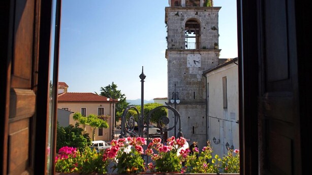 Visão através de uma porta aberta de um edifício com uma torre de relógio, um poste de luz e flores em flor em primeiro plano.