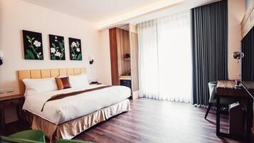 Quarto de hotel com uma cama grande, uma mesa e uma cadeira. Há três quadros na parede e uma janela com cortinas.