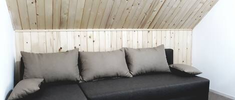 Quarto com teto de madeira inclinado, um sofá escuro com várias almofadas e um pequeno alto-falante preto no lado direito.