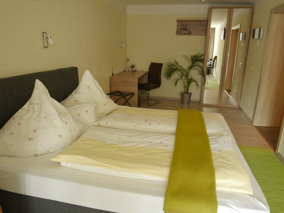 Quarto de hotel com uma cama, escrivaninha, cadeira e planta em vaso.