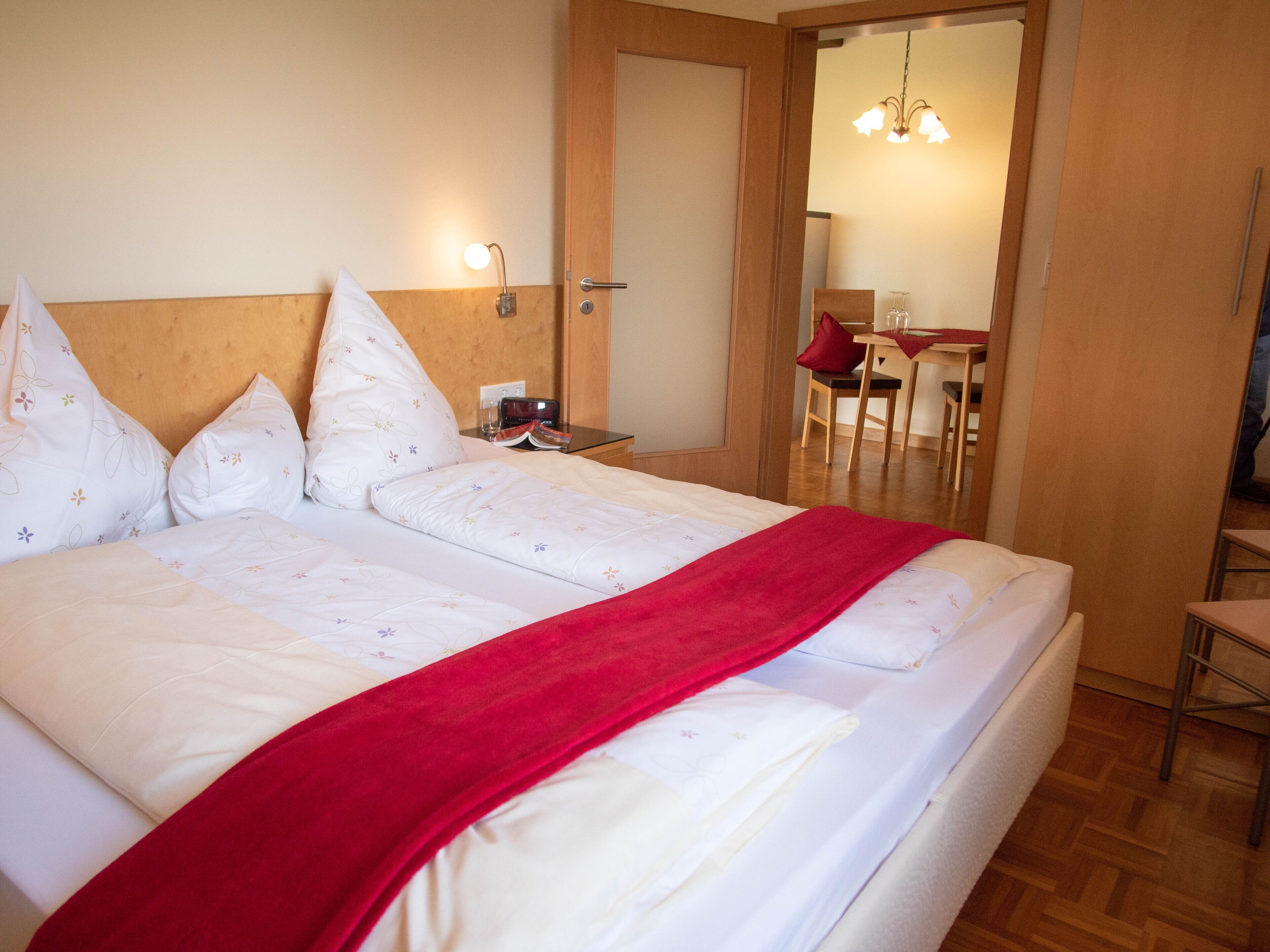 Cama bem arrumada com lençóis brancos e manta vermelha, mesa de cabeceira com abajur e porta de madeira que dá para uma sala de jantar.