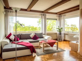 Uma sala de estar moderna com sofá, mesa de centro e abajur. Janelas amplas com cortinas, um radiador e um vaso de planta.