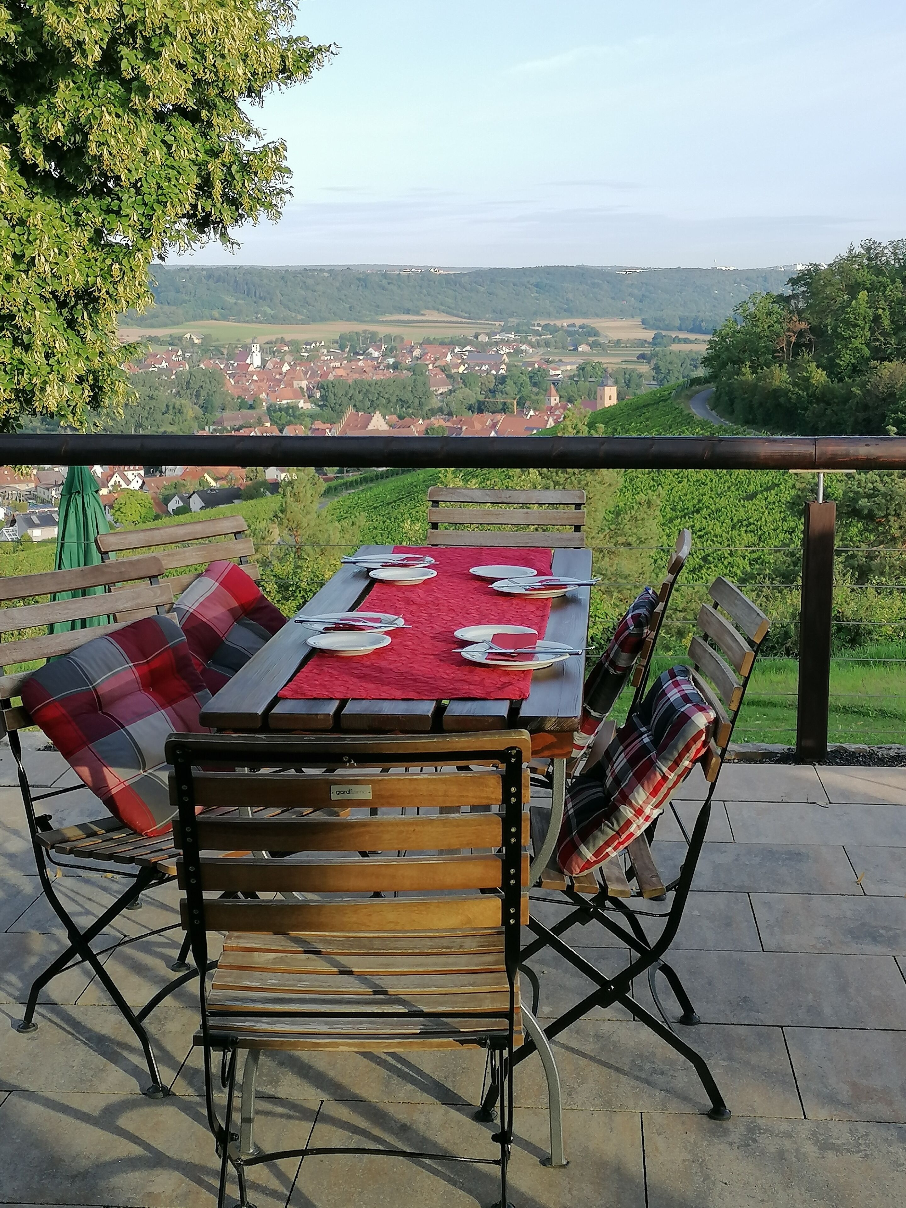Área de refeições ao ar livre com uma mesa posta para quatro pessoas, rodeada por cadeiras com almofadas xadrez, com vista para uma paisagem pitoresca de uma aldeia e colinas.