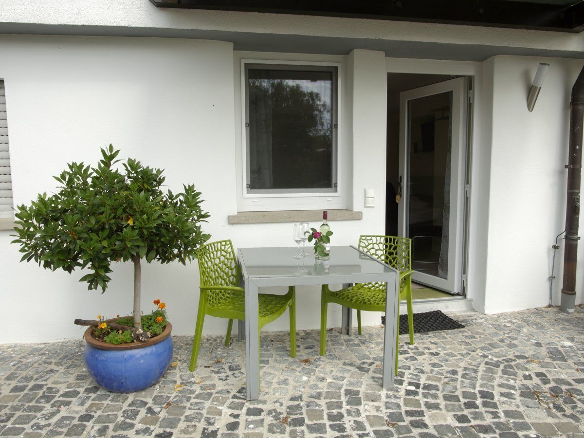 Um pátio externo com uma mesa, cadeiras, uma árvore em vaso e uma porta que dá para uma área coberta.