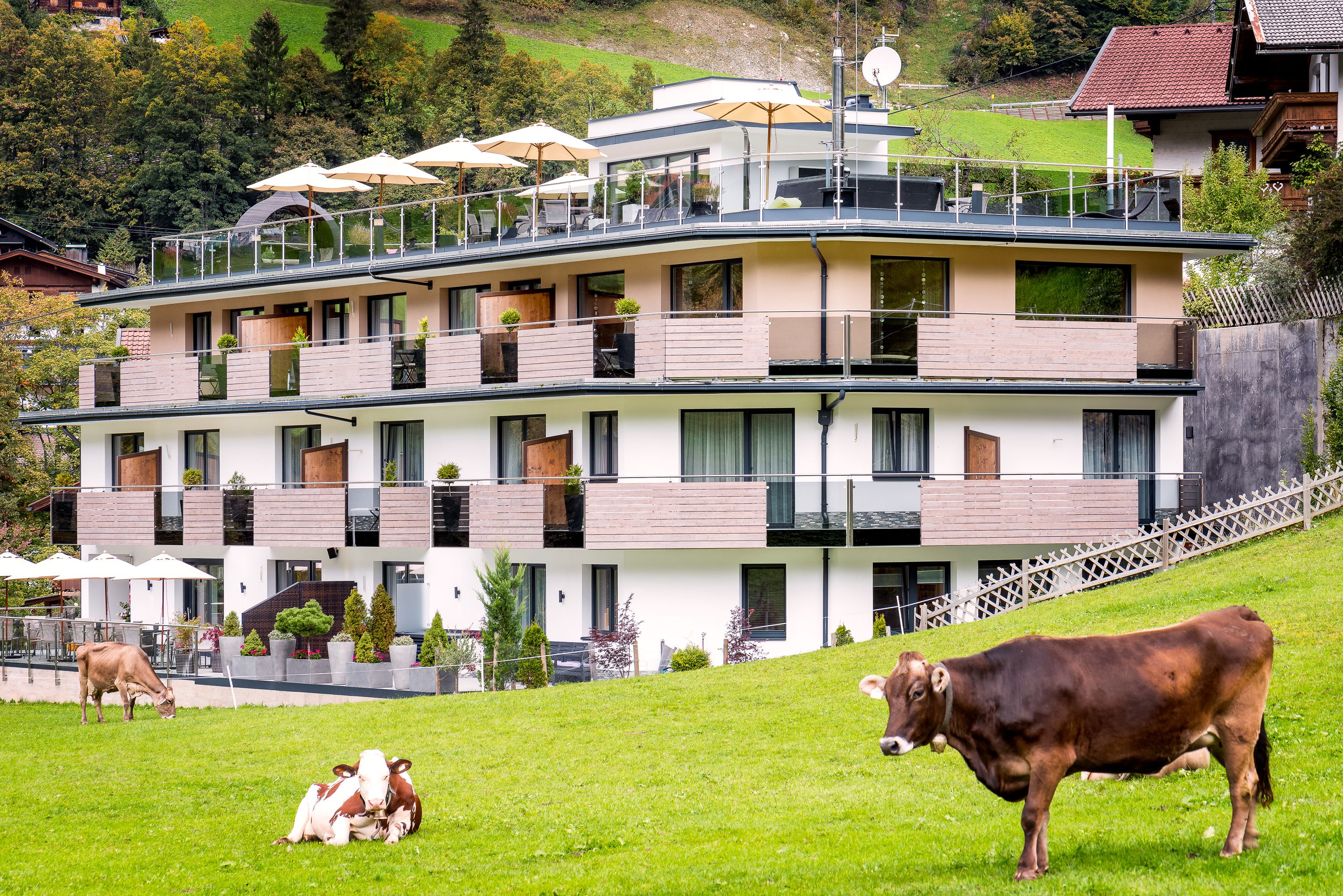 Um hotel com vários andares, varandas, área externa com mesas e cadeiras e uma área gramada com vacas.