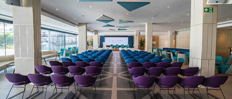 Sala de conferências com fileiras de cadeiras roxas, um palco com uma tela e uma janela grande com vista para o exterior.