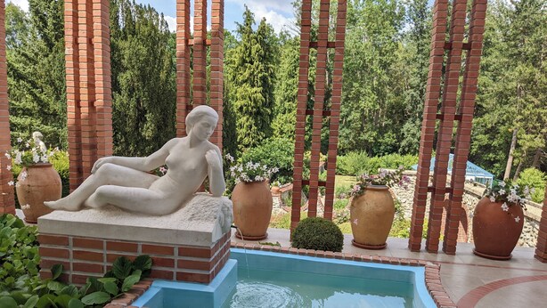 Escultura de uma figura reclinada em um jardim com piscina, cercada por construções de tijolo e plantas em vasos.
