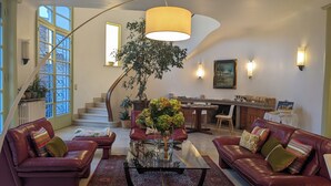 Uma sala de estar ampla com sofás de couro, uma mesa de centro de vidro e uma planta grande. Uma escada com corrimão curvo leva a um andar superior.