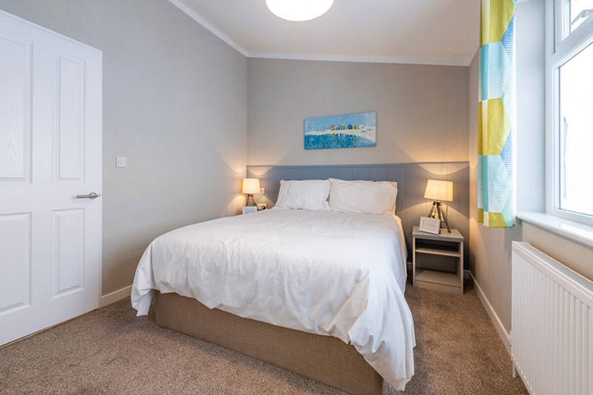 Quarto com cama, duas mesas de cabeceira com luminárias, uma janela com cortinas e um quadro na parede.
