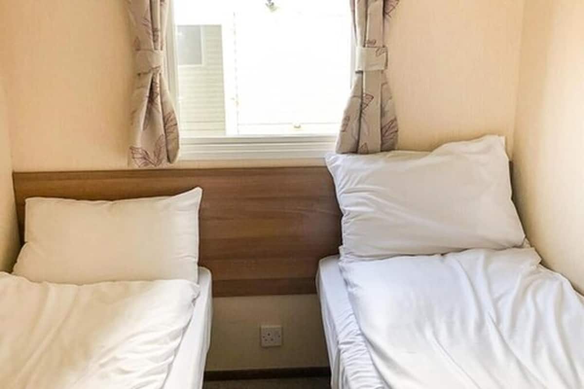 Acomodação simples, com um quarto único, duas camas de solteiro, cabeceira de madeira e uma janela com cortinas.