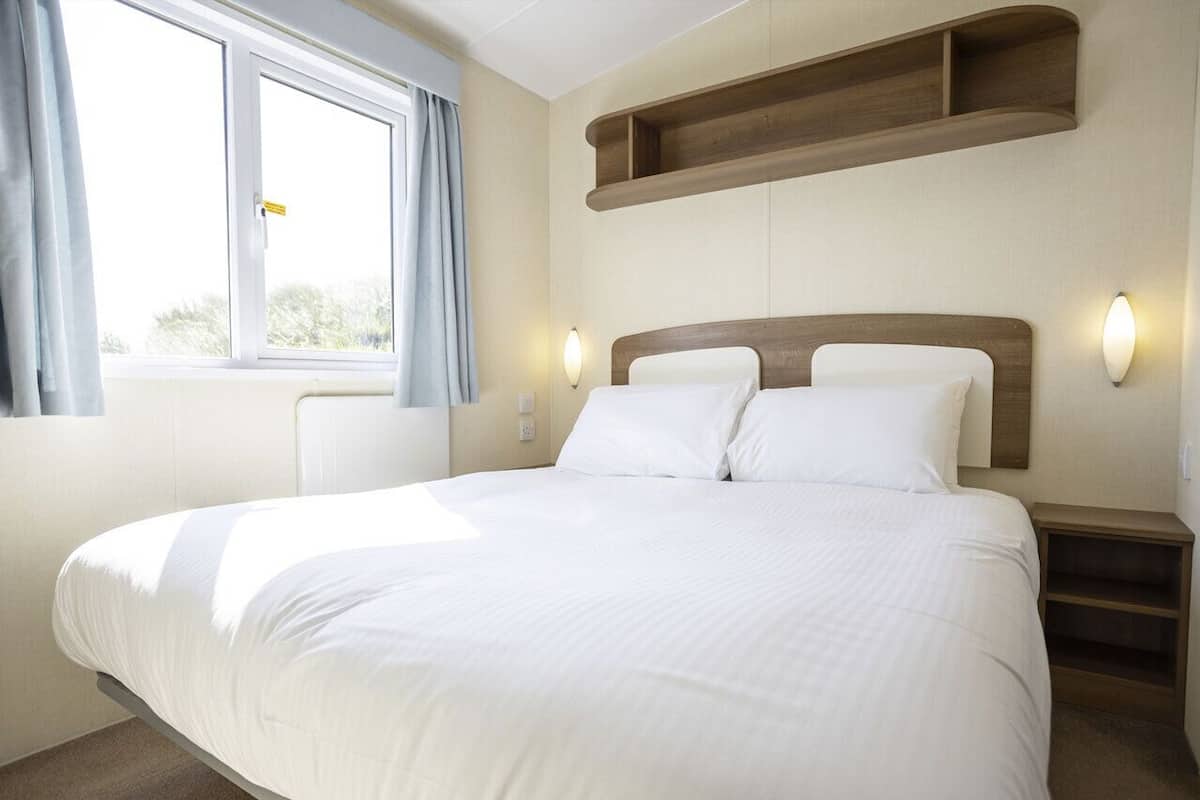 Quarto de hotel com uma cama grande, janela com cortinas, mesa de cabeceira e luminárias fixadas na parede.