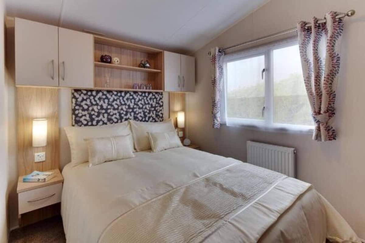 Quarto com cama, mesa de cabeceira, guarda-roupa e janela com cortinas.