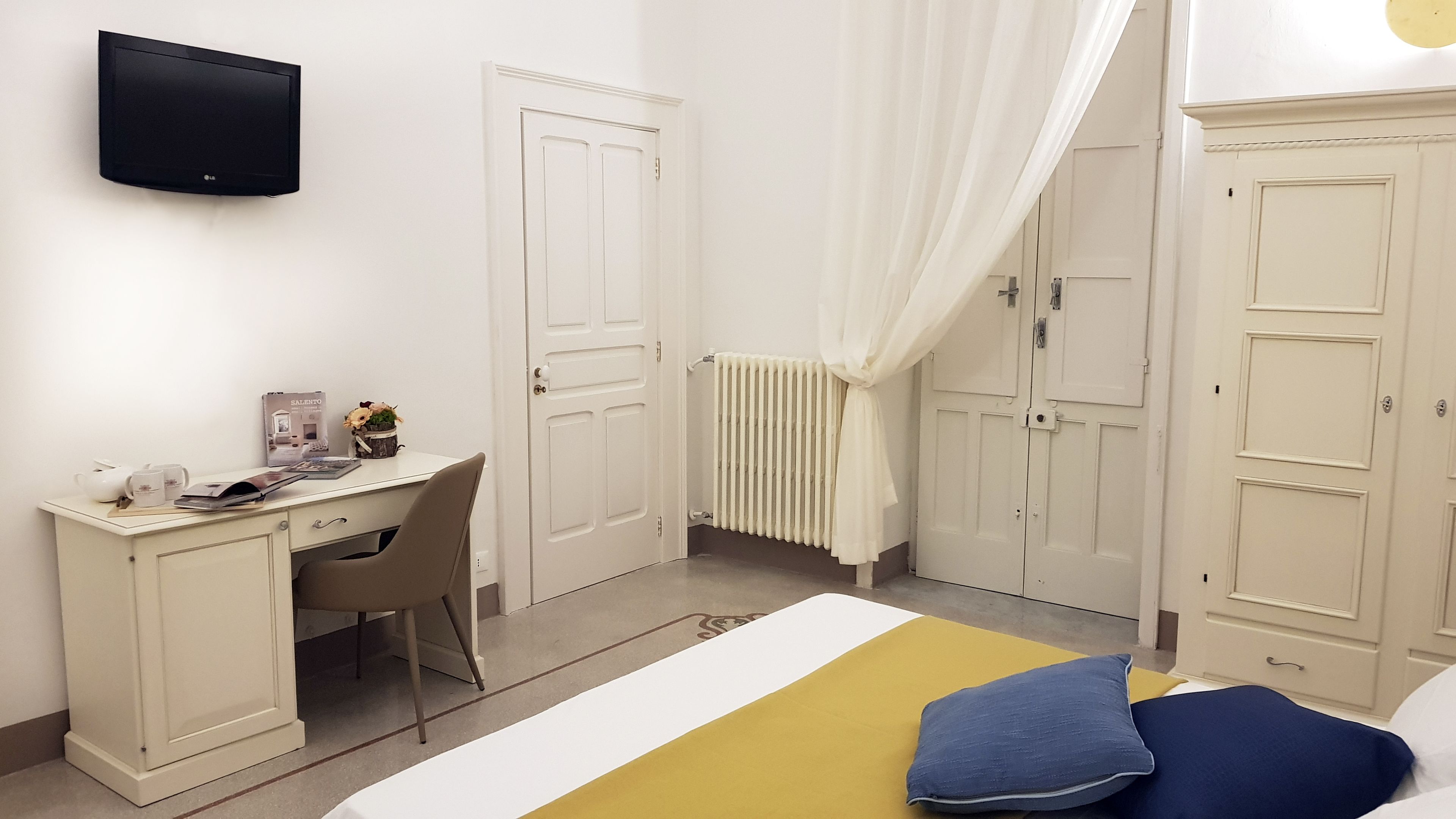 Quarto com cama, escrivaninha, cadeira, radiador e televisão na parede.