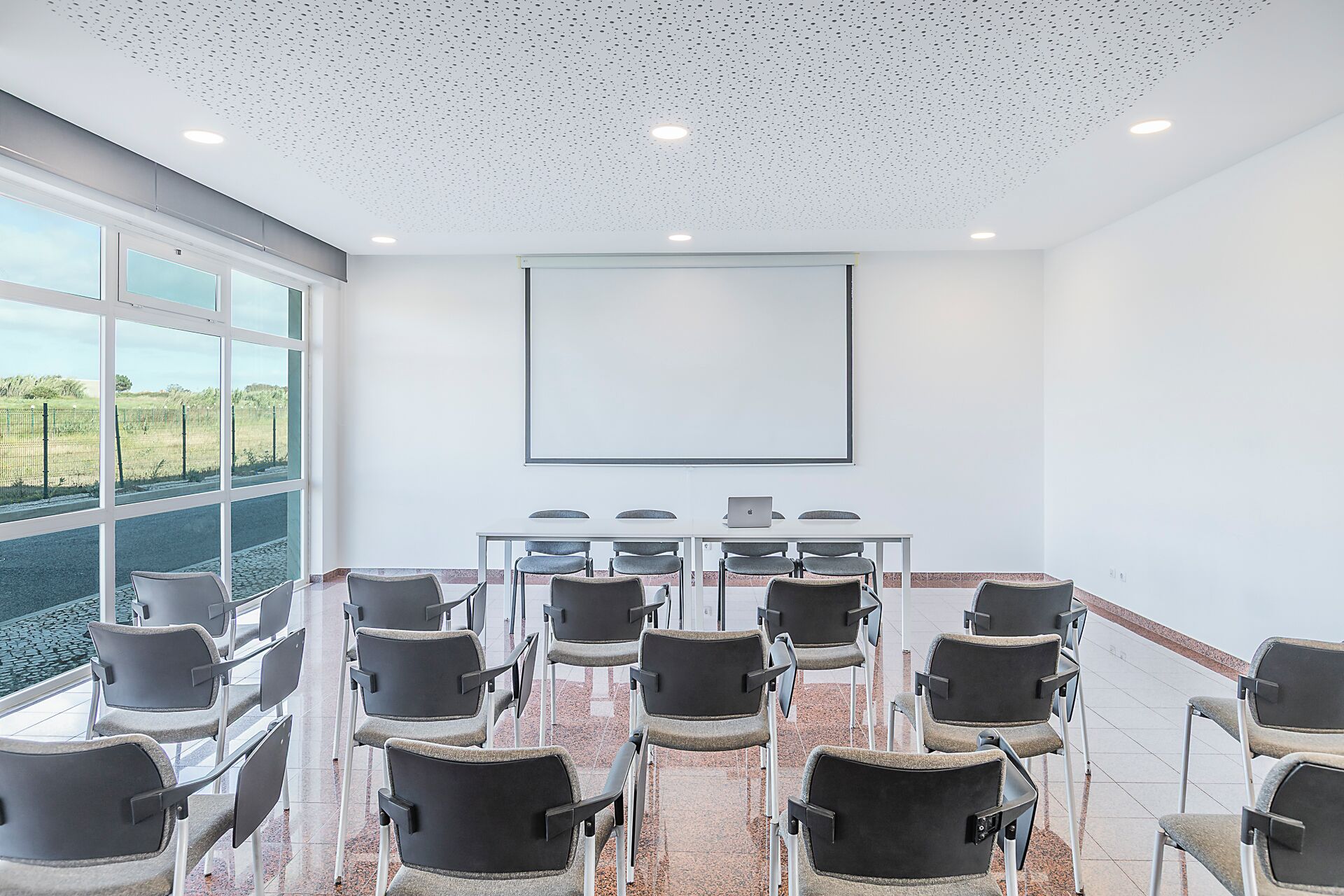 Uma sala de conferências com uma janela grande, uma tela de projetor e fileiras de cadeiras.