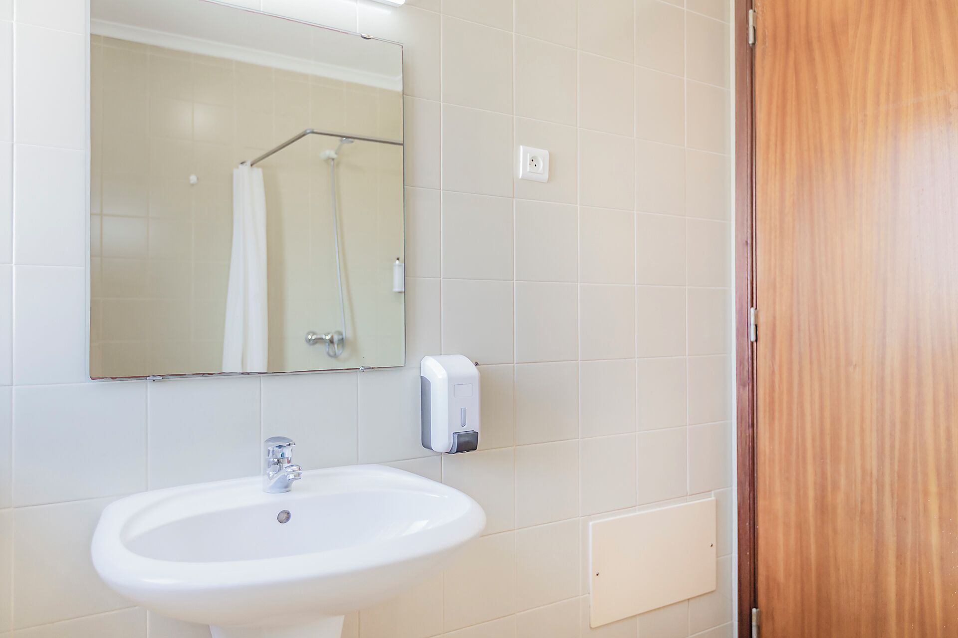 Uma casa de banho com uma pia branca, um espelho, uma cabine de duche e um dispensador de sabão.