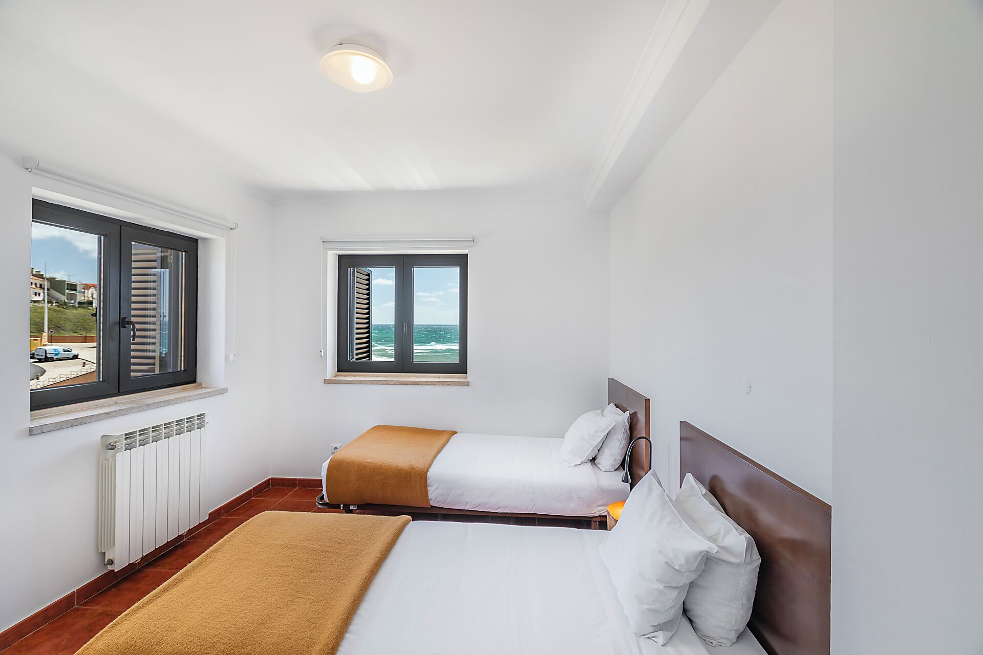 Quarto com cama, janela vista para o mar, radiador e luz no teto.