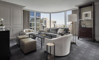 Quarto de hotel moderno com área de estar curva, uma mesa de centro e vista para a paisagem urbana através de grandes janelas.