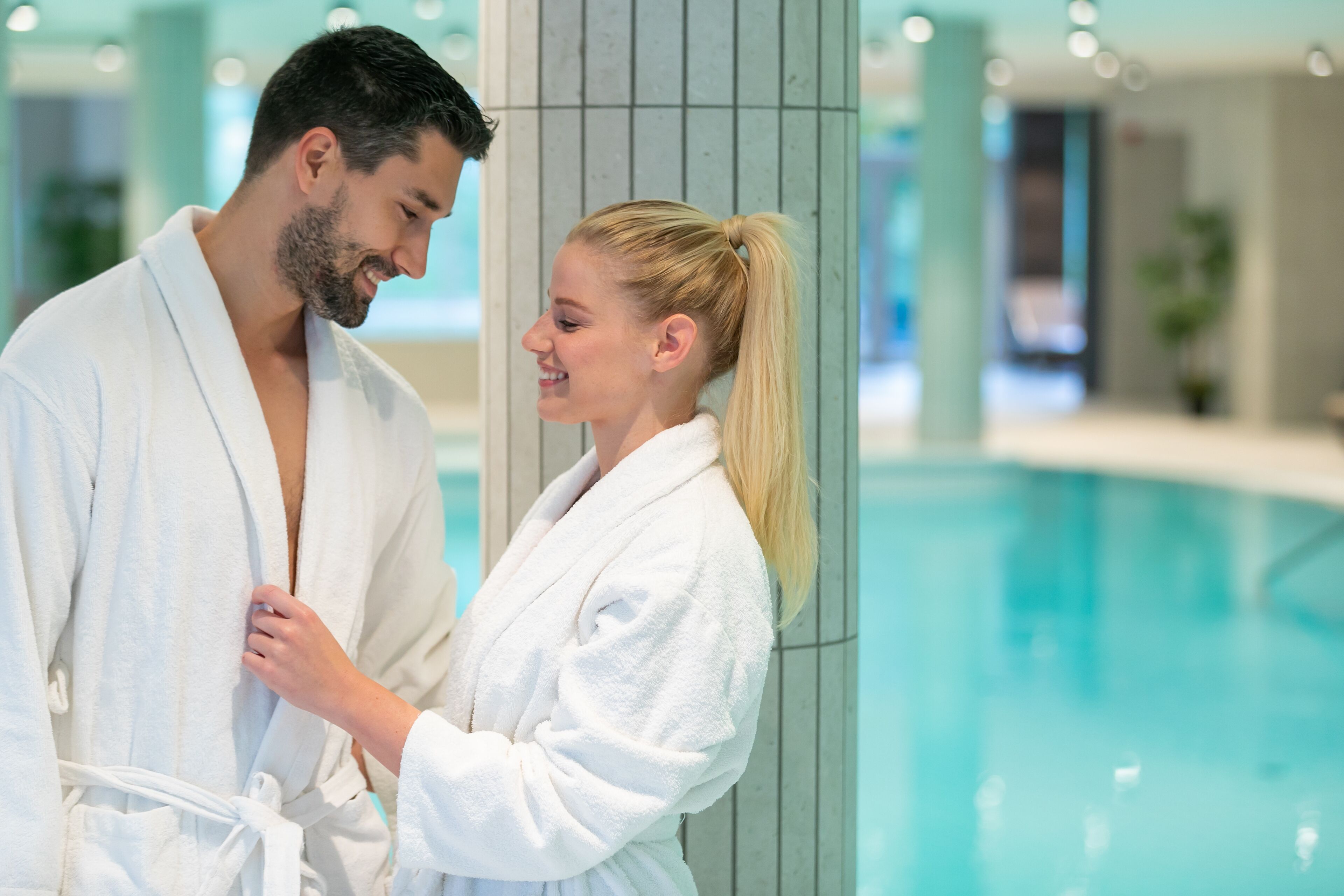 Um casal em roupões brancos ao lado de uma piscina com parede azulejada e iluminação interna.