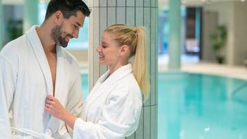 Um casal em roupões brancos ao lado de uma piscina com parede azulejada e iluminação interna.