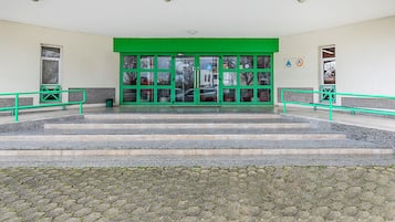 Entrada de um edifício com porta verde, degraus de acesso e área pavimentada em frente.