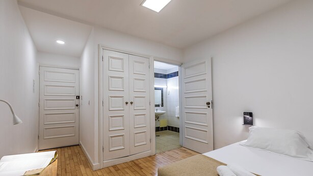 Quarto de hotel com cama, uma escrivaninha com abajur e um banheiro visível através de uma porta aberta.