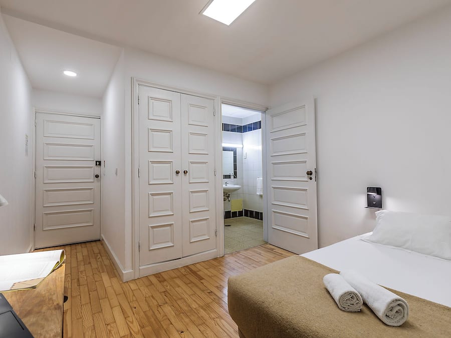 Quarto de hotel com cama, uma escrivaninha com abajur e um banheiro visível através de uma porta aberta.