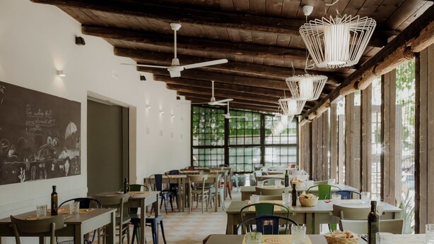 Restaurante com vigas de madeira no teto, luminárias pendentes brancas e piso xadrez. Mesas montadas com cadeiras, copos e cardápio.