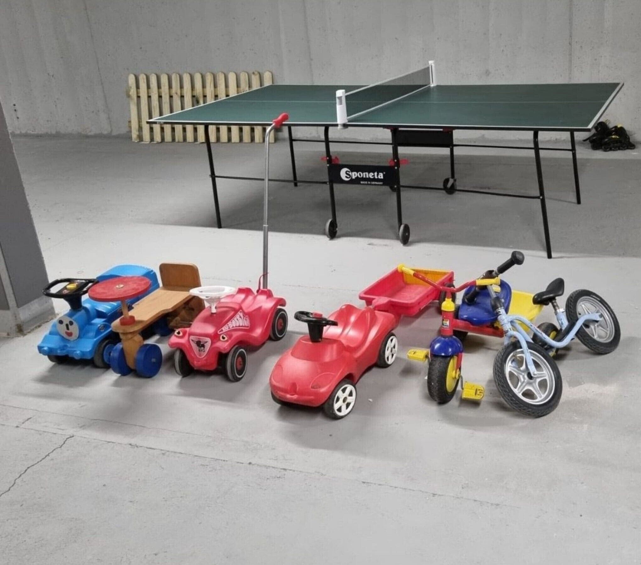 Diversos brinquedos infantis, incluindo carrinhos e uma triciclo, dispostos sobre um piso de concreto, com uma mesa de tênis de mesa ao fundo.