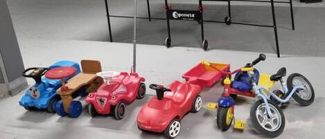Diversos brinquedos infantis, incluindo carrinhos e uma triciclo, dispostos sobre um piso de concreto, com uma mesa de tênis de mesa ao fundo.