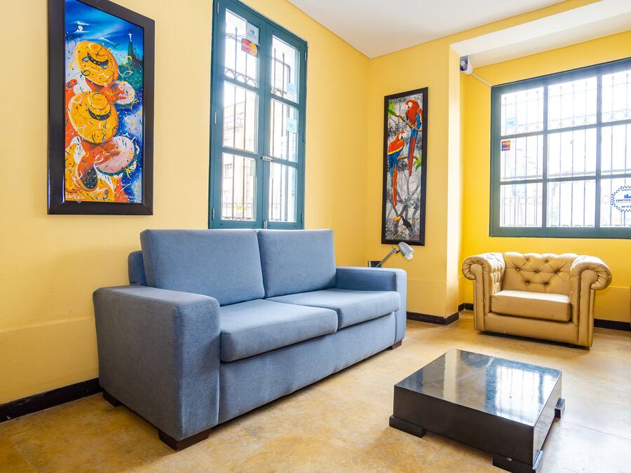 Uma sala de estar com um sofá azul, uma poltrona bege e uma mesa de centro. Há duas pinturas nas paredes e uma janela com vista para o exterior.