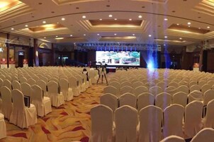 Sala de conferências com fileiras de cadeiras brancas, um palco com uma tela grande e piso coberto de carpete.