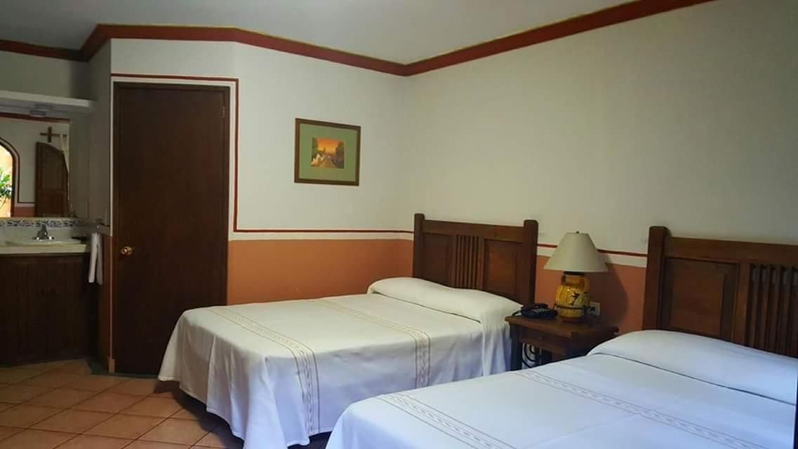 Quarto de hotel com duas camas de solteiro, cabeceira de madeira, mesa de cabeceira com abajur, porta e pequeno quadro na parede.