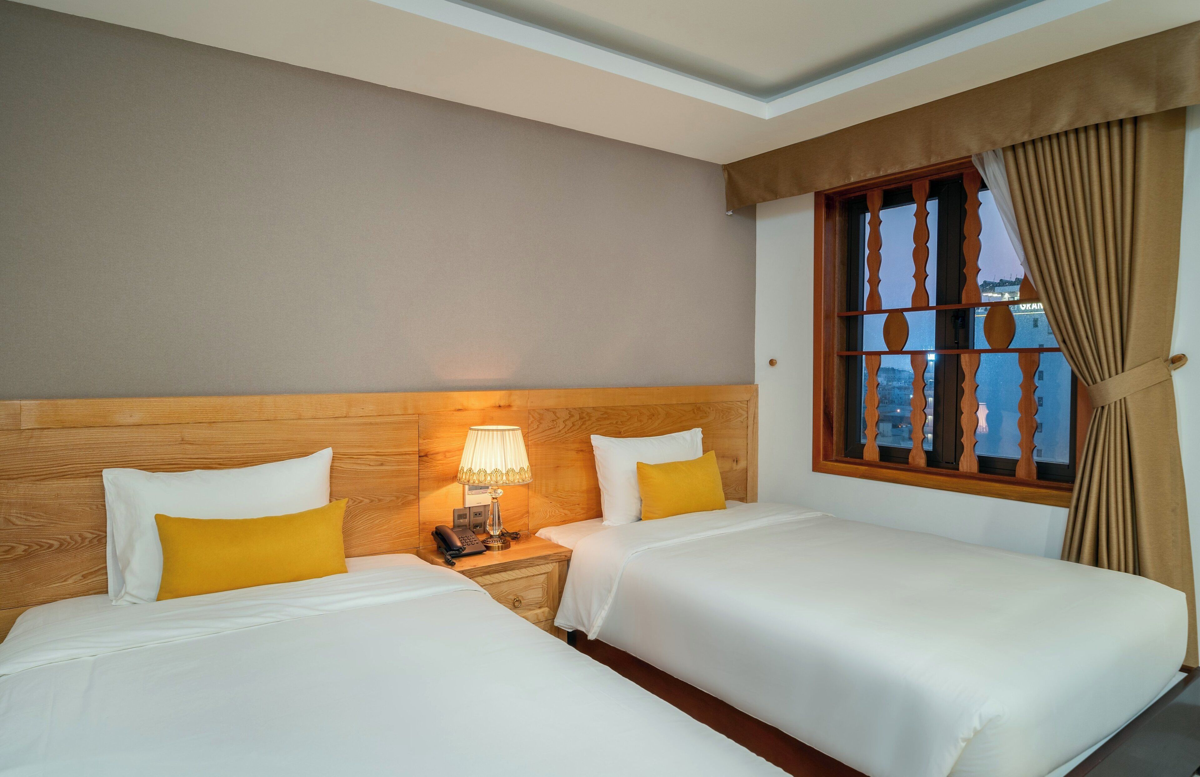 Quarto de hotel com duas camas, cabeceira de madeira, mesa de cabeceira com abajur e janela com cortinas.