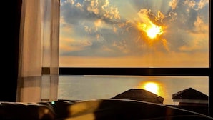 Uma cama com vista para o pôr do sol no mar, uma janela com cortinas translúcidas e um pequeno edifício ao longe.