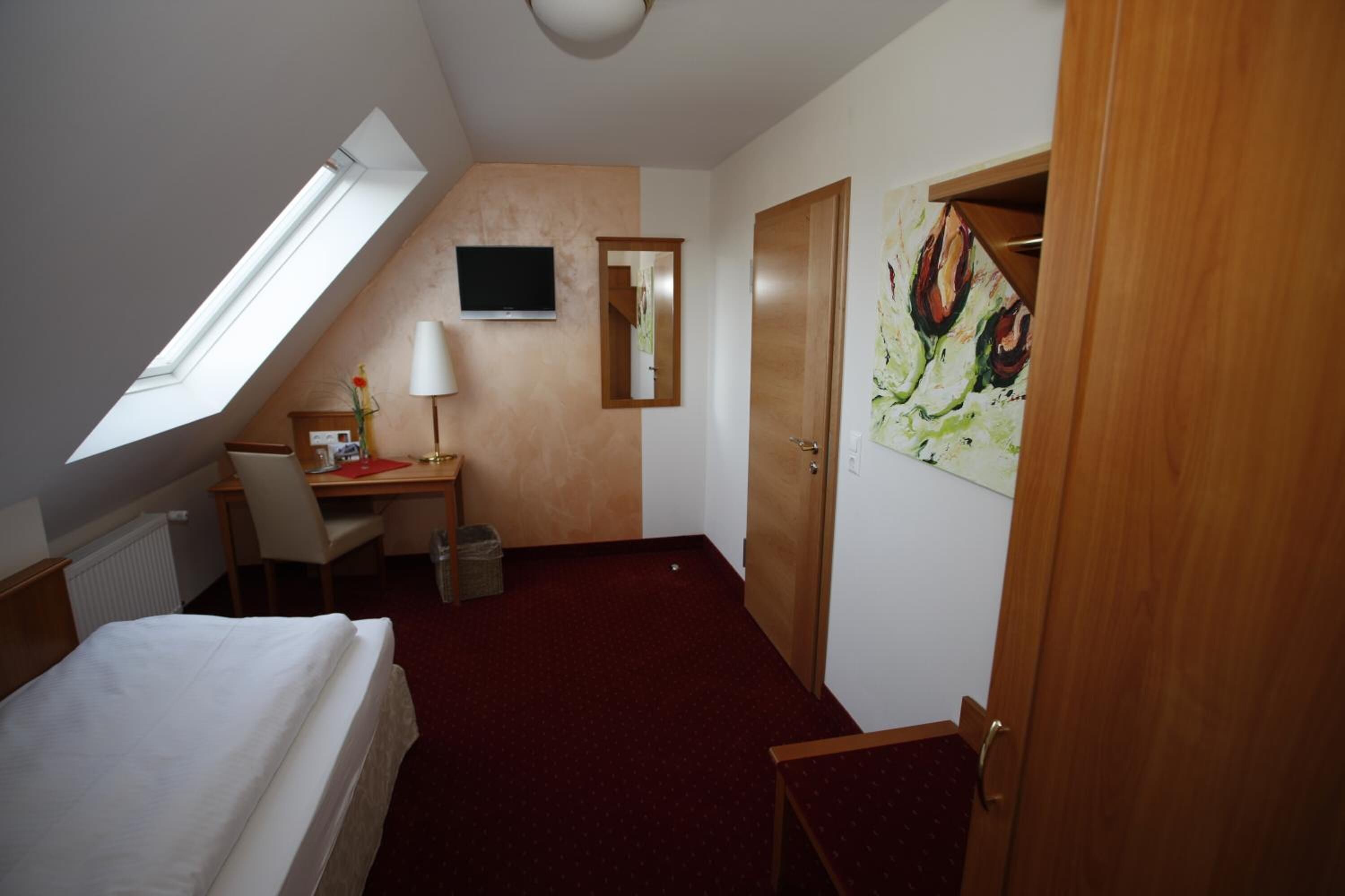 Quarto de hotel com cama, escrivaninha, cadeira, televisão, espelho e porta.