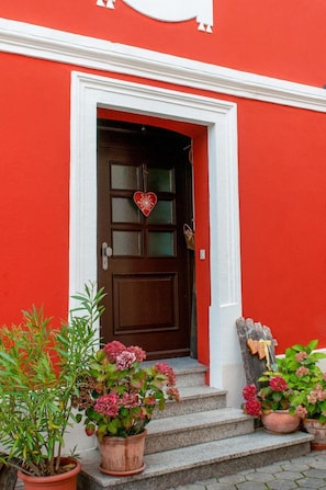 Um edifício vermelho com porta branca, plantas em vasos e uma decoração em formato de coração na porta.