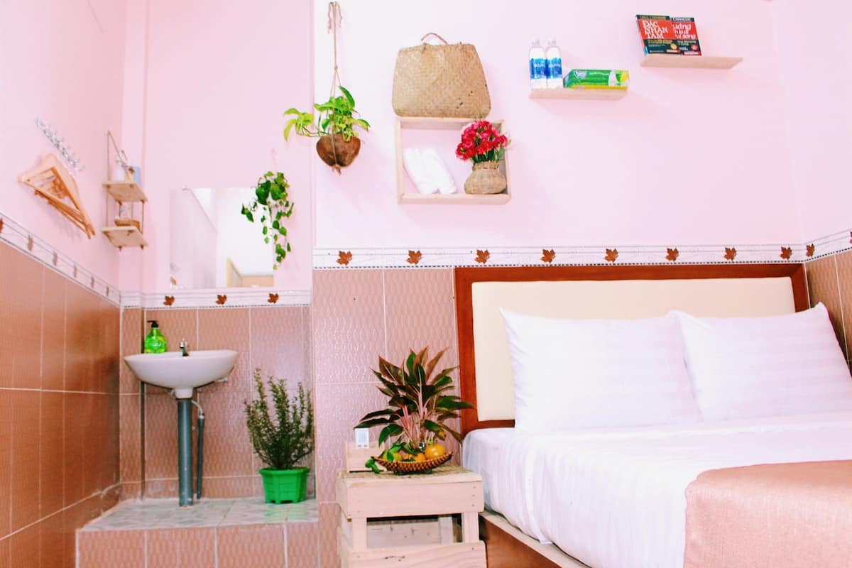 Quarto de hotel em tons de rosa, com uma cama, uma mesa de cabeceira com uma planta, uma planta pendente, uma pequena prateleira com artigos de higiene pessoal e um espelho.