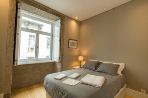 Quarto com cama, mesa de cabeceira, abajur, quadro na parede e janela com vista para um edifício.
