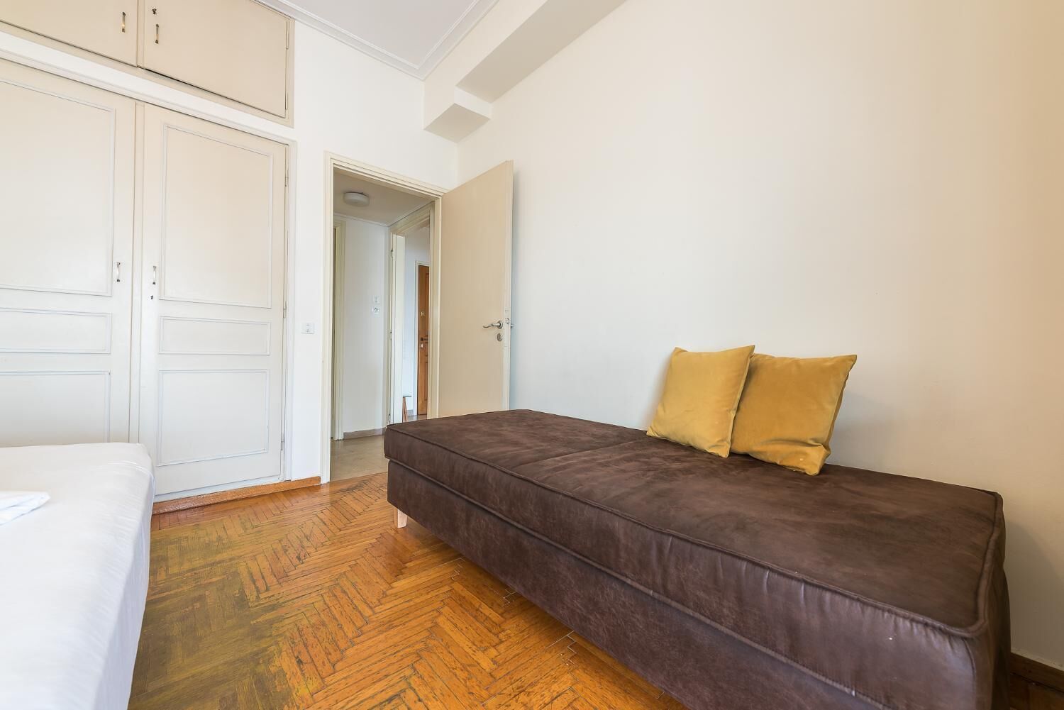 Quarto com sofá-cama marrom, duas almofadas, cama branca e guarda-roupa.