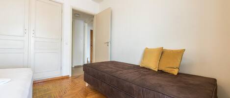 Quarto com sofá-cama marrom, duas almofadas, cama branca e guarda-roupa.