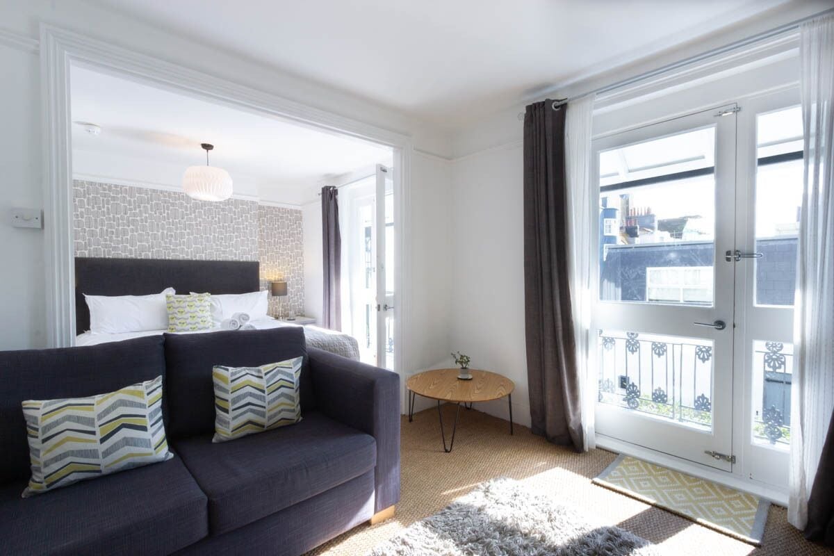 Quarto de hotel moderno com sofá escuro, cama com roupa de cama branca, mesa redonda pequena e varanda com vista.