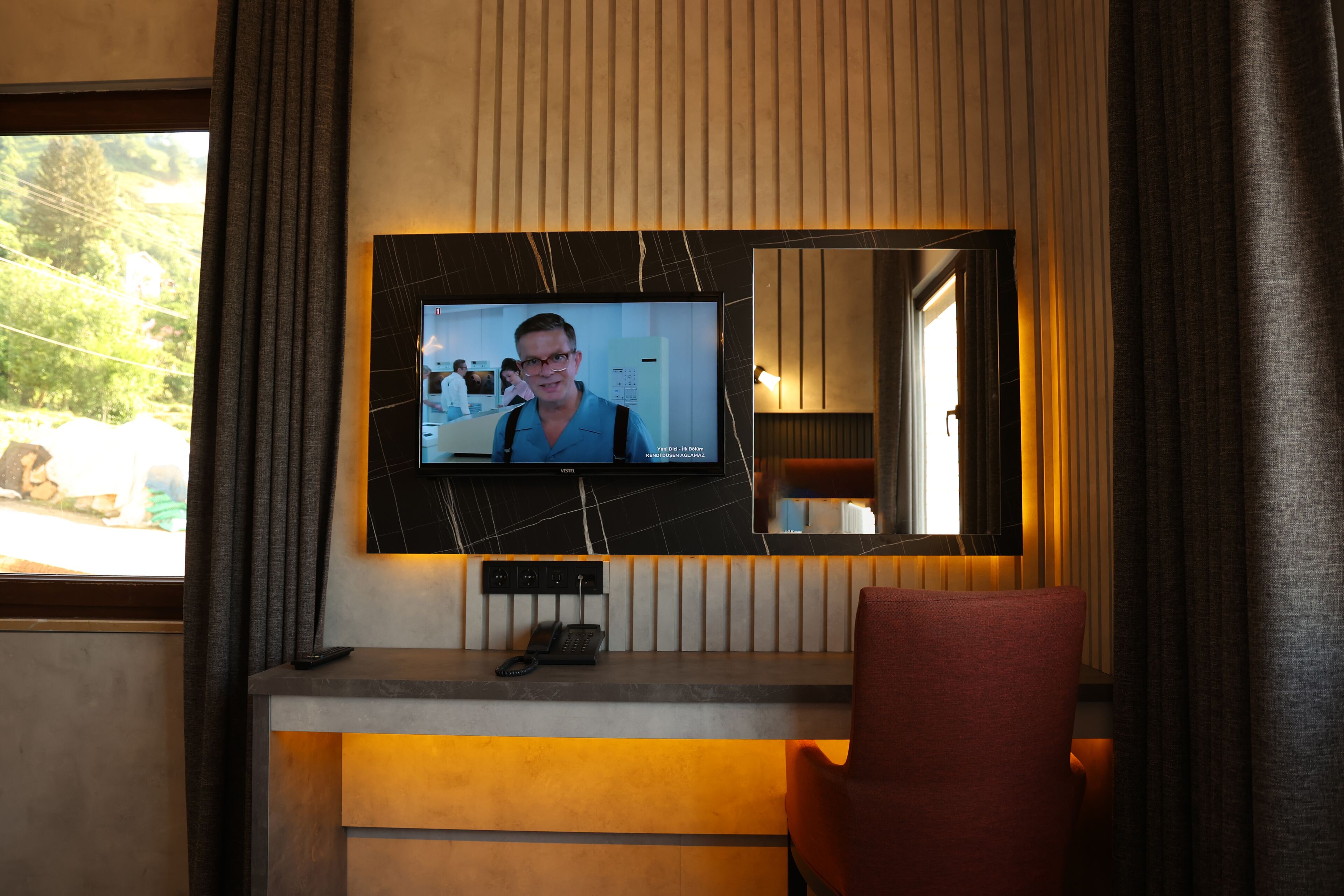 Quarto de hotel com uma televisão de tela grande fixada na parede, uma mesa com telefone e uma cadeira vermelha.