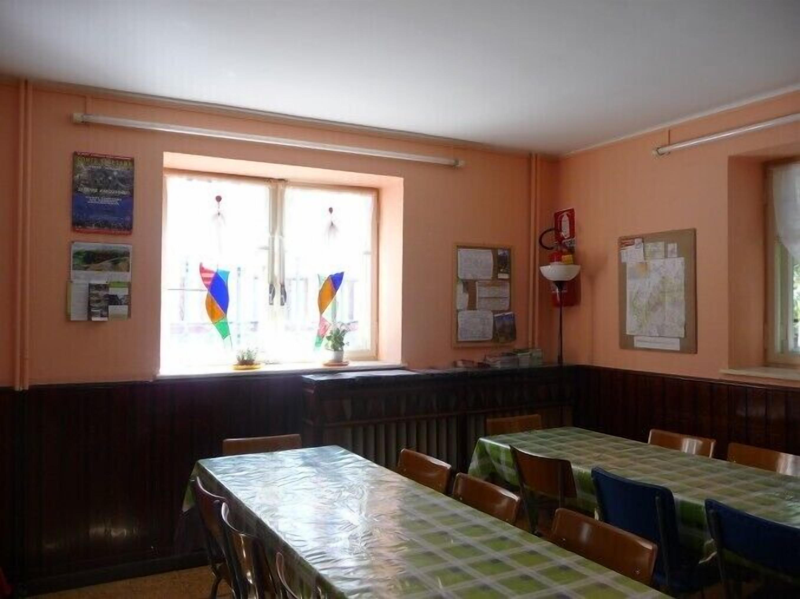 Uma sala de jantar com mesas cobertas por toalhas verdes, uma janela adornada com bandeiras coloridas e painéis de avisos nas paredes.